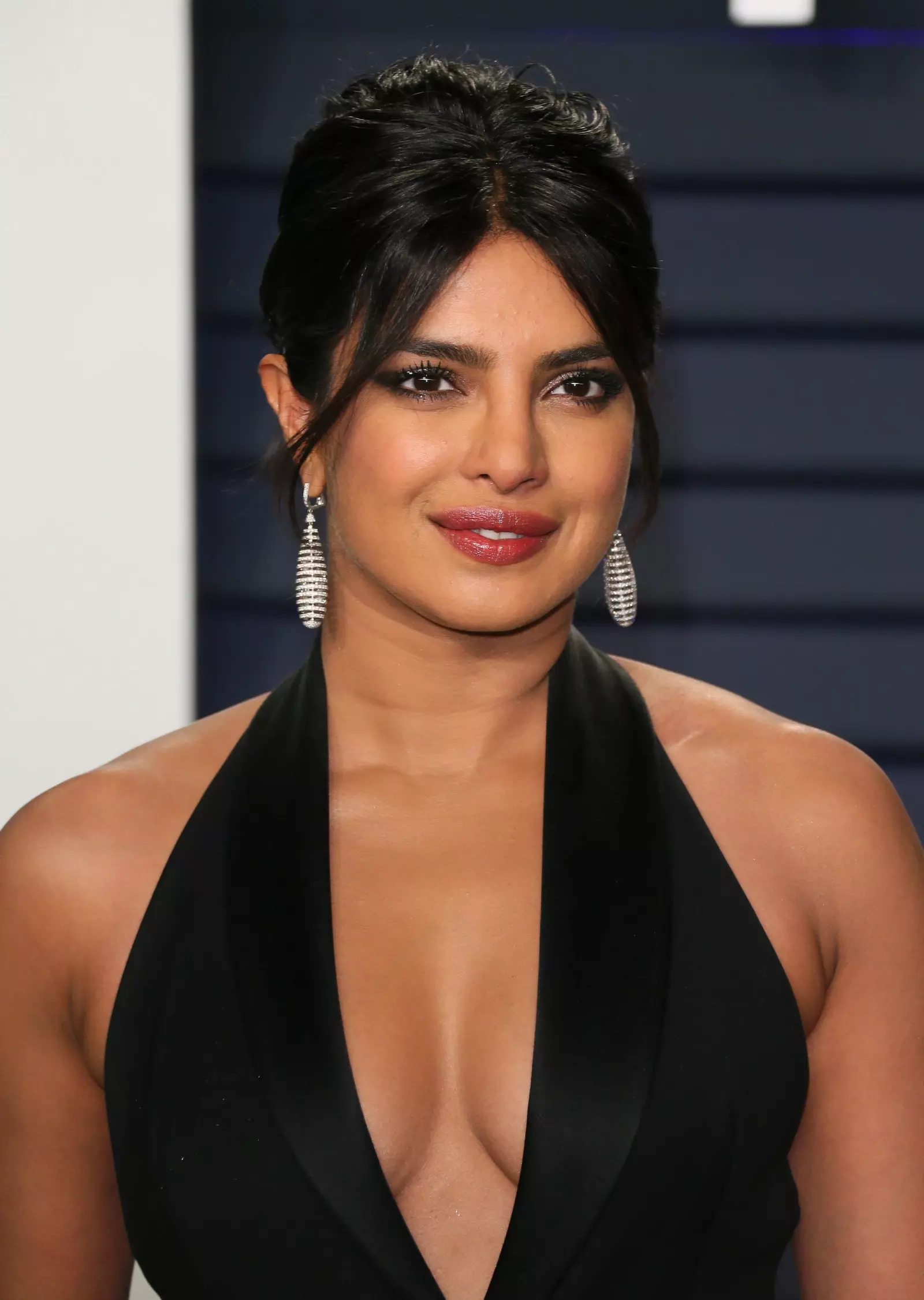 Priyanka Chopra ina seasamh ar an gcairpéad dearg agus a cuid gruaige in updo ag caitheamh cluaise airgid dangly agus dubh gearrtha íseal ...